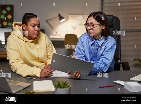 Portrait Of Two Young Women Discussing Documentation While Working Late At Night In Office Stock
