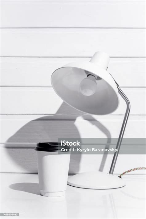 검은 뚜껑으로 닫힌 커피가 든 종이 컵과 흰색 테이블 위에 흰색 램프가 서 있고 흰색 나무 배경이 있습니다 0명에 대한 스톡