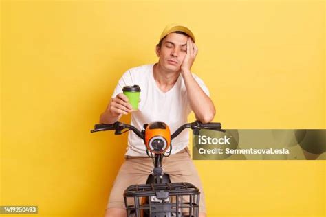 하얀 티셔츠와 모자를 쓰고 전기 스쿠터를 타고 졸린 느낌 커피 컵을 들고 눈을 감고 노란 배경에 고립 된 지루한 피곤한 남자의 실내 샷 남자에 대한 스톡 사진 및 기타