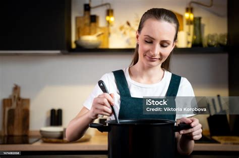 앞치마를 입은 만족스럽고 행복한 젊은 주부 여자는 수프 냄비 숟가락을 들고 냄비에서 수프를 맛본다 가정에있는 사람들의 생활