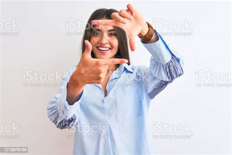 행복한 얼굴로 손과 손가락으로 프레임을 만드는 고립 된 흰색 배경 위에 서있는 파란색 우아한 셔츠를 입은 젊은 아름다운 여성 창의력과 사진 개념 몸짓에 대한 스톡 사진 및
