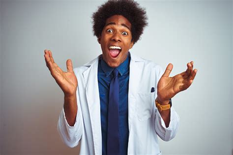 고립된 흰색 배경 위에 코트를 입은 젊은 아프리카 계 미국인 의사가 미친 듯이 축하하고 팔을 들고 눈을 뜨고 흥분한 채 성공을