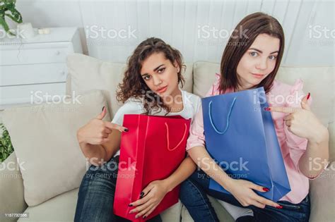 소파에 앉아 미소를 지으며 카메라를 바라보며 매우 즐겁고 빨간색과 파란색 상점 가방을 껴안고있는 두 명의 쇼핑 중독자 소녀 표정에 만족하는 얼굴 표정 2명에 대한 스톡 사진