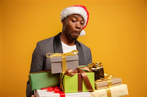 재킷과 크리스마스 모자를 쓴 어두운 피부의 남자가 그의 얼굴에 놀란 표정으로 손에 선물 상자 더미를 바라보고 있습니다 12월에 대한 스톡 사진 및 기타 이미지 Istock
