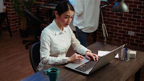 Smiling Asian Employee Writing Important Emails At Computer Desk Next To Full Mug Of Coffee