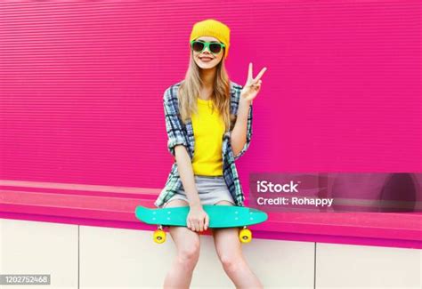 분홍색 배경에 화려한 노란색 모자를 쓰고 평화 표지판 제스처를 보여주는 스케이트 보드와 멋진 웃는 여자 T 셔츠에 대한 스톡 사진 및 기타 이미지 Istock