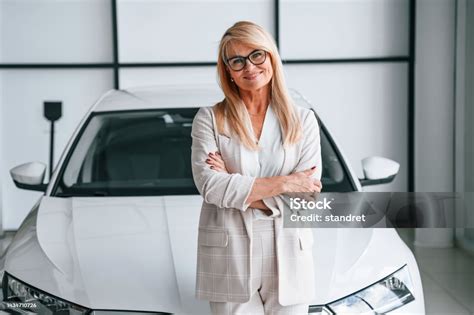 팔짱을 끼고 서 있습니다 흰색 정장을 입은 여자가 자동차 대리점에 있다 자동차 대리점에 대한 스톡 사진 및 기타 이미지