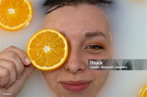 우유와 오렌지가 들어간 욕조에 누워 있는 여자가 아름답게 웃고 있다 여자는 손에 주황색 원을 들고 한쪽 눈을 가리고 있습니다 평면도 세로화 목욕 우유와 오렌지 감귤에 대한