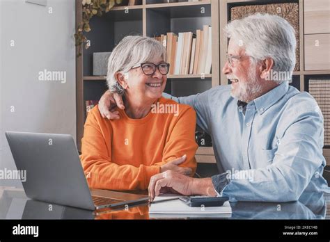 Happy Elderly 60s Couple Sit Rest On Couch At Home Pay Household Expenses Online On Computer