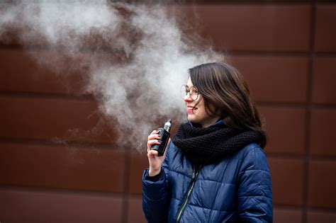 Vape 십대 겨울에 거리에서 현대 갈색 배경 맞은 편에 전자 담배를 흡연 파란색 재킷과 안경에 젊은 예쁜 흰색 백인 소녀 나쁜 습관 십대에 대한 스톡 사진 및 기타 이미지
