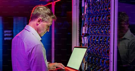 Serious Mature Network Engineer Standing At Open Server Cabinet And Using Laptop While