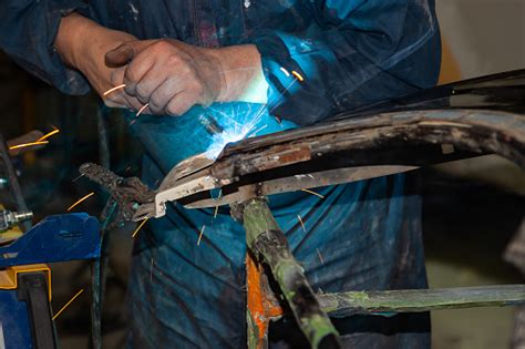 클로즈업 보기 강한 남자는 팔에 장갑 없이 작업 바지에 용접기입니다 금속 제품은 차고 워크샵에서 용접 기계로 용접 블루 연기와 오렌지 스파크는 측면에 비행 강철에 대한 스톡