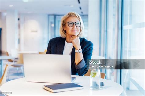 50대의 자신감 넘치는 성숙한 사업가가 창문을 바라보며 꿈꾸고 중년의 경험 많은 고위 여성 전문가가 열린 공간 사무실에서 노트북을 작업하고 있다 원격으로 일하는 여성 기업가