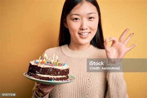 노란 고립 된 배경에 불타는 촛불로 생일 케이크를 들고 있는 젊은 아시아 여성은 손가락으로 확인 기호를하고 우수한 기호 개념에