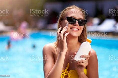 금발 여성은 뺨에 자외선 차단제 태양 크림을 바르고 수영장 근처의 얼굴에 바릅니다 웃는 예쁜 소녀는 스파의 피부에 플라스틱 병에서 선탠 크림을 넣습니다 선탠 로션을 곁들인