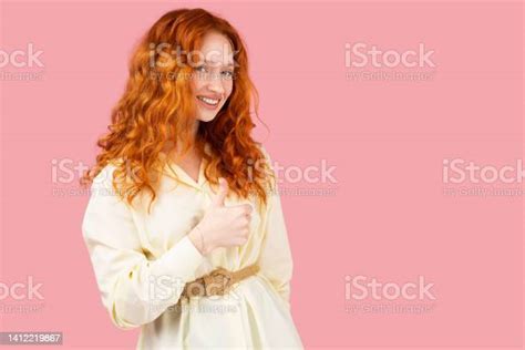 미소 짓는 빨간 머리 젊은 여성이 분홍색 배경과 복사 공간에 좋은 표시로 엄지 손가락을 보여주는 카메라를보고 개성 개념에 대한