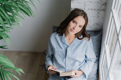 편안한 쾌적한 찾고 여자의 상단보기는 캐주얼 국내 정장을 입고 좋은 빛과 비전을 위해 창틀에 앉아 독서 책을 즐기고 외로움과 여가 시간을 즐긴다 취미 개념 20 24세에