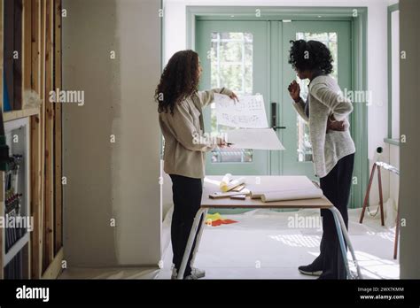 Side View Of Client And Female Architect Comparing Blueprints While Standing Near Table At Under