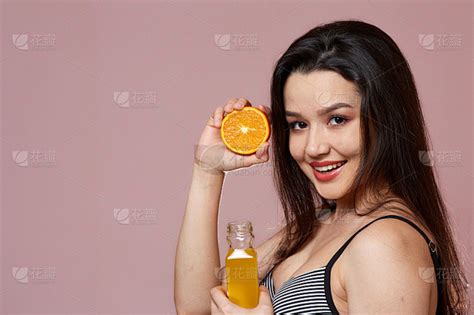 年轻美丽苗条的亚洲女孩穿着泳衣手里拿着新鲜的果汁和桔子微笑在粉红色孤立的背景上