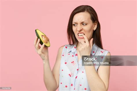 여름 옷에 관련 된 젊은 여성은 분홍색 파스텔 벽 배경에 고립 된 신선한 잘 익은 녹색 아보카도 과일을 들고 손톱을 갉 아 먹는
