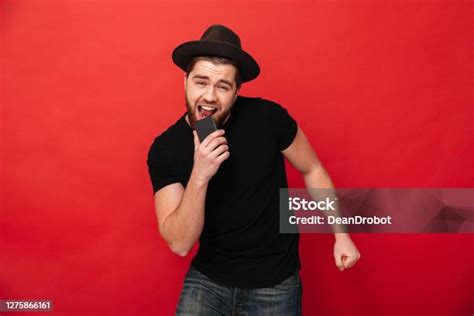 캐주얼하고 검은 모자를 쓴 수염을 기쁜 남자의 이미지와 빨간색 벽 위에 고립 된 마이크와 같은 휴대 전화를 들고 듣는 것 T 셔츠에 대한 스톡 사진 및 기타 이미지 Istock