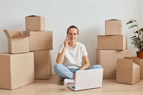 개인 더미가 달린 골판지 상자 근처 바닥에 앉아 카메라를 보고 스마트폰을 통해 이야기하고 컴퓨터에서 온라인으로 작업하고 움직이는 흰색 티셔츠를 입은 긍정적인 여성 30 34