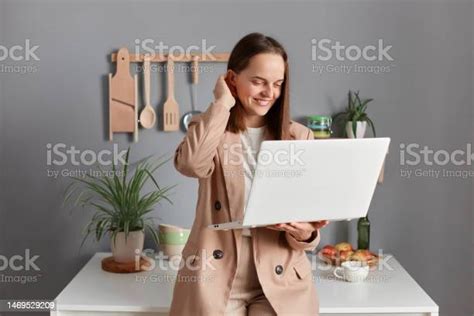 베이지색 정장을 입은 아름다운 만족스러운 갈색 머리 여성이 집 부엌 테이블 근처에 서서 노트북으로 작업하고 미소를 지으며 디스플레이를 바라보는 실내 사진 30 34세에 대한