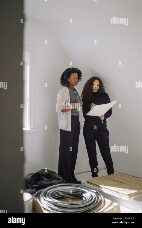 Female Architect And Client Discussing Over Blueprint While Standing In Attic At Under