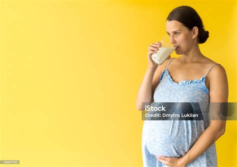꽃 무늬 블루 드레스를 입은 초상화 귀여운 임신 한 젊은 예쁜 여성은 노란 배경에 배를 문질러 우유 한 잔을 들고 있습니다 모성 여성스러움 건강한 식습관 유제품 여름 컨셉