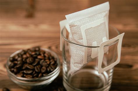 집에서 커피를 만드는 추세 갈색 배경에 커피 원두가 든 유리잔에 신선한 음료가 담긴 가방을 드립한다 컵에 양조하기위한 분쇄 커피 용지 필터를 사용하여 커피를 드립합니다