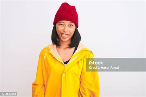 젊은 아름 다운 중국 여자 비옷과 양모 모자 위에 고립 된 흰색 배경 튀어 나와 혀를 밖으로 재미 있는 표정으로 행복 감정 개념입니다 가을에 대한 스톡 사진 및 기타 이미지