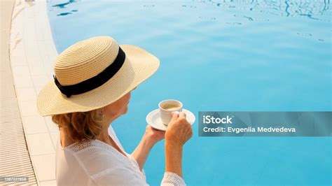 흰색 드레스를 입은 밀짚 모자를 쓰고 수영장에서 아침 식사를 하는 50세 여성 커피 한 잔과 함께 수영장 에 앉아 여자 좋은 아침과 낮 계획 건강한 생활방식에 대한 스톡