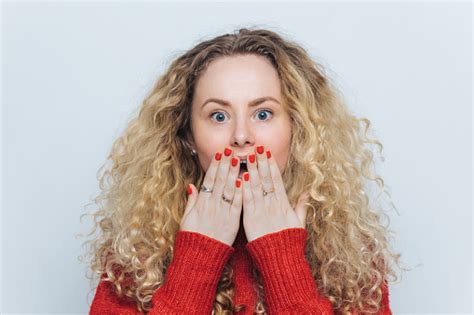 예기치 않은 표정으로 감정적 인 사랑스러운 여성은 손으로 입을 덮고 흰색 스튜디오 배경 위에 고립 된 빨간 매니큐어 곱슬 머리를 가지고 있습니다 충격 젊은 귀여운 여성