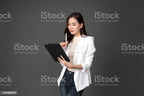 긴 소매에 흰색 코트를 입은 여성 임원 스튜디오에서 회색 배경을 배경으로 사진을 찍고 있다 그녀는 노트북 태블릿과 펜을 들고 있습니다 직장에 서서 열심히 생각합니다 25