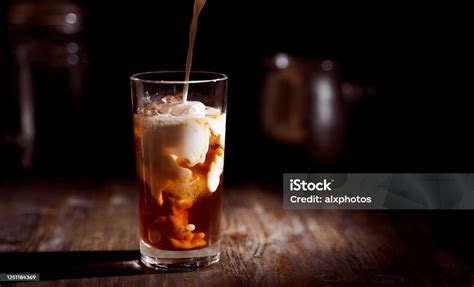 크림이 쏟아진 높은 유리에 아이스 커피를 쏟아 붓고 오래된 소박한 나무 테이블에 얼음 조각과 콩이 들어옵니다 검은 색 배경에 튜브와 차가운 음료 0명에 대한 스톡 사진 및
