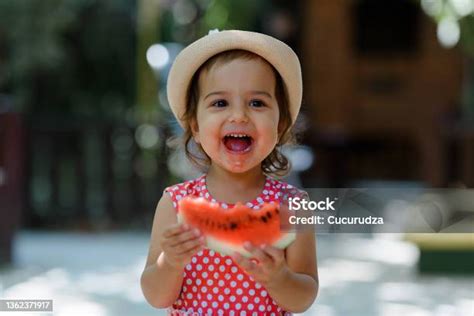 흰색 물방울 무늬와 모자가있는 빨간 드레스를 입은 어린 2 살 소녀는 수박의 매우 재미있는 조각을 먹고 있습니다 개념에 대한 스톡 사진 및 기타 이미지 Istock