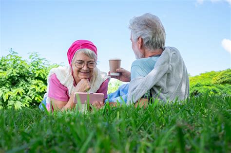 공공 공원의 잔디밭에 누워있는 노인 미소 짓는 커플 휴대 전화를 보는 여자 커피 컵을 들고 있는 남자의 초상화 자유 시간과 은퇴를 즐기는 낭만적 인 현대 커플 2명에 대한