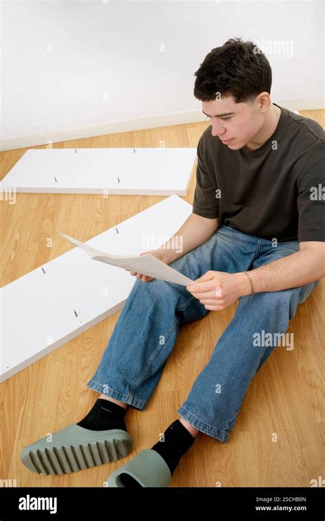 A Young Man Sits On A Wooden Floor Reading Assembly Instructions For New Furniture Parts Spread