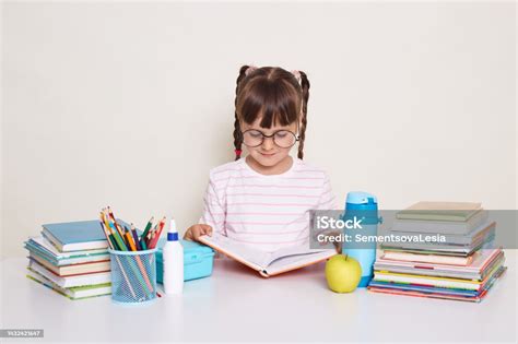 어두운 머리카락과 머리띠를 가진 진지한 세심한 어린 여학생이 책과 독서로 둘러싸인 테이블에 앉아 수업 중에 문학 과제를 수행하는 이미지 6 7 살에 대한 스톡 사진 및 기타
