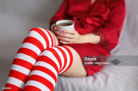 빨간 흰색 무릎 양말과 실크 나이티를 입은 여성이 손에 찻잔을 들고 소파에 앉아 있습니다 가정 생활에 대한 스톡 사진 및 기타