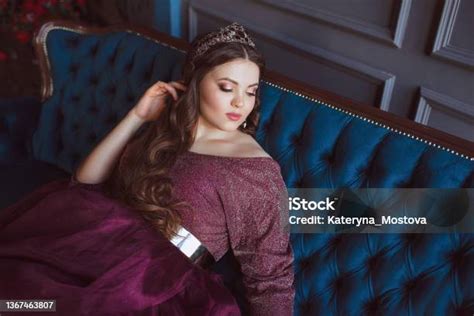 긴 보라색 여왕의 드레스와 크라운에 아름다운 젊은 예쁜 귀여운 여자 공주 의 초상화 긴 머리와 파란색 벨벳 소파에 앉아 메이크업 왕족에 대한 스톡 사진 및 기타 이미지