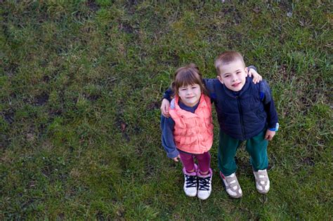 녹색 잔디에 서있는 캐주얼 한 옷을 입은 두 명의 귀여운 아이들의 위에서 보면 어깨 위로 서로 껴안고 수줍은 미소를 지으며 행복한 어린 시절과 우정 개념 감정에 대한 스톡