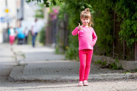 분홍색 캐주얼 의류와 긴 금발 조랑말 꼬리에 귀여운 작은 소녀의 전체 길이 초상화 흐리게 밝은 햇볕이 잘 드는 거리 배경에 엄지 손가락 제스처와 함께 카메라에 미소 어린