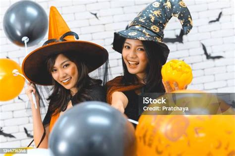 할로윈 파티를위한 검은 종이 박쥐 모양으로 장식 된 흰색 벽돌 배경에 풍선을 들고 마녀 할로윈 의상을 입은 두 명의 아름다운 젊은 아시아 여성 2명에 대한 스톡 사진 및