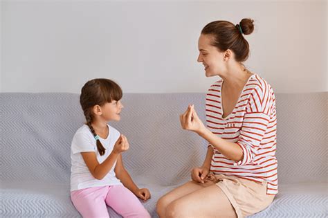 회색 소파에 앉아있는 동안 귀여운 아이 소녀와 음성 결함에 종사하는 여성 전문 물리 치료사의 측면 보기 말하기를 개선하기위한 언어 수업을 가진 머리띠가있는 아이 25 29