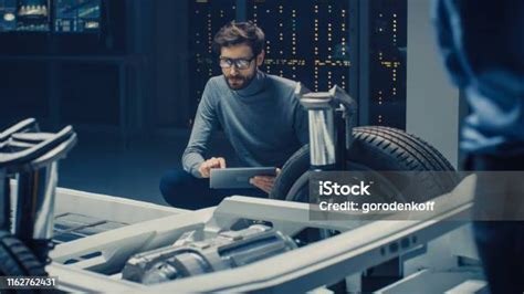 하이브리드 전기 자동차 섀시 플랫폼 프로토 타입 옆에 앉아 자동차 디자인 엔지니어 디자인 향상을위한 태블릿 컴퓨터를 사용하여 서스펜션 휠 엔진 및 배터리가 있는 차량