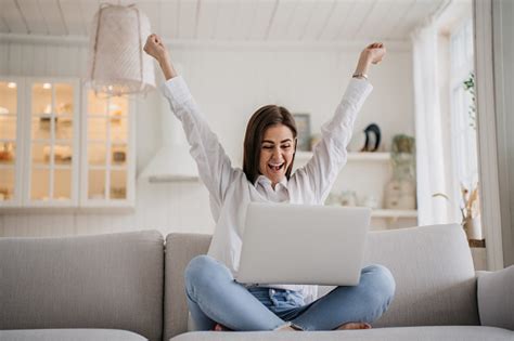 부엌 소파에 캐주얼하게 앉아 노트북을 들고 승자 표정으로 손을 들고 새로운 직업을 갖게 되어 기뻐하는 젊은 놀란 여성 좋은 소식을 받고 축하하는 소녀 행복한 사람들의