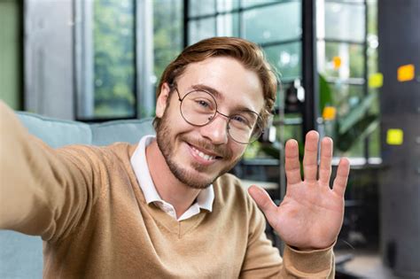 클로즈업 사진 젊은 남성 블로거 학생 사업가가 손을 흔들고 인사하고 카메라와 이야기합니다 사무실 책상에 앉아 웃으며 사진을 찍는다 남성에 대한 스톡 사진 및 기타 이미지