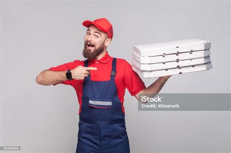 파란색 유니폼을 입고 수염과 젊은 행복 배달 남자와 빨간 T셔츠 서 들고 가리키는 회색 배경에 골 판지 피자 상자 더미를 손가락