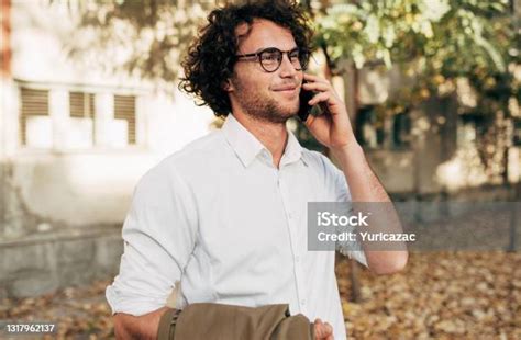 거리에서 휴대 전화로 말하는 똑똑한 젊은 남자의 야외 촬영 흰색 셔츠와 안경에 곱슬 머리를 가진 잘 생긴 남성은 도시 거리에서 자신의 휴대 전화에 전화를 걸고 20 29세에